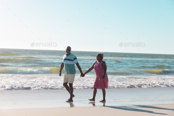 Happy african american senior couple holding hands and walking at seashore under clear sky Stock ...