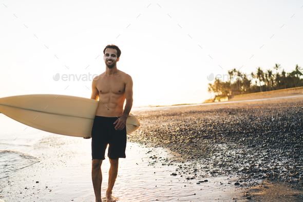 Cheerful male surfer walking on beach with board under arm Stock Photo ...