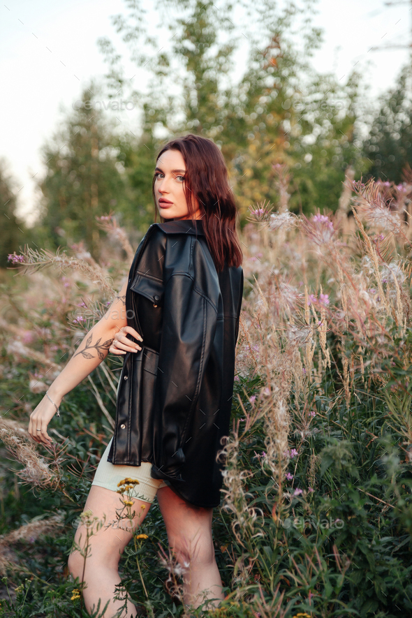 Pensive jewish young woman model in leather jacket posing at greenery ...