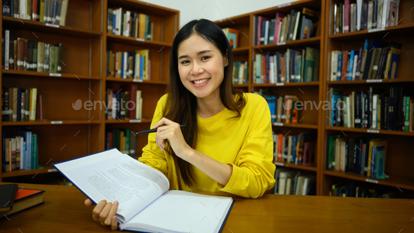 Smiling female student sitting among bookshelves in library reading ...