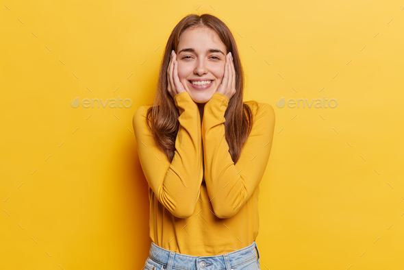 Beautiful cheerful woman smiles toothily keeps both hands on cheeks ...