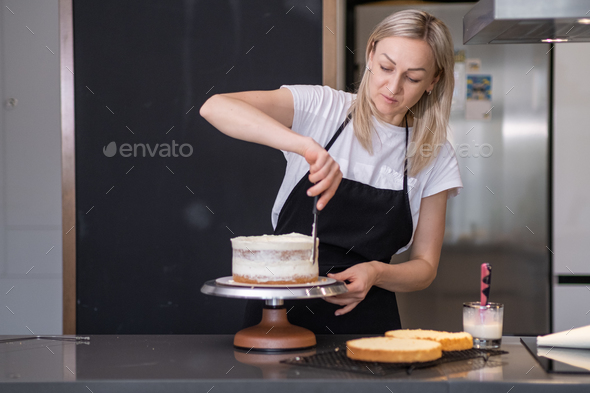 Concentrated blonde woman smearing white cream around fresh cake layers ...