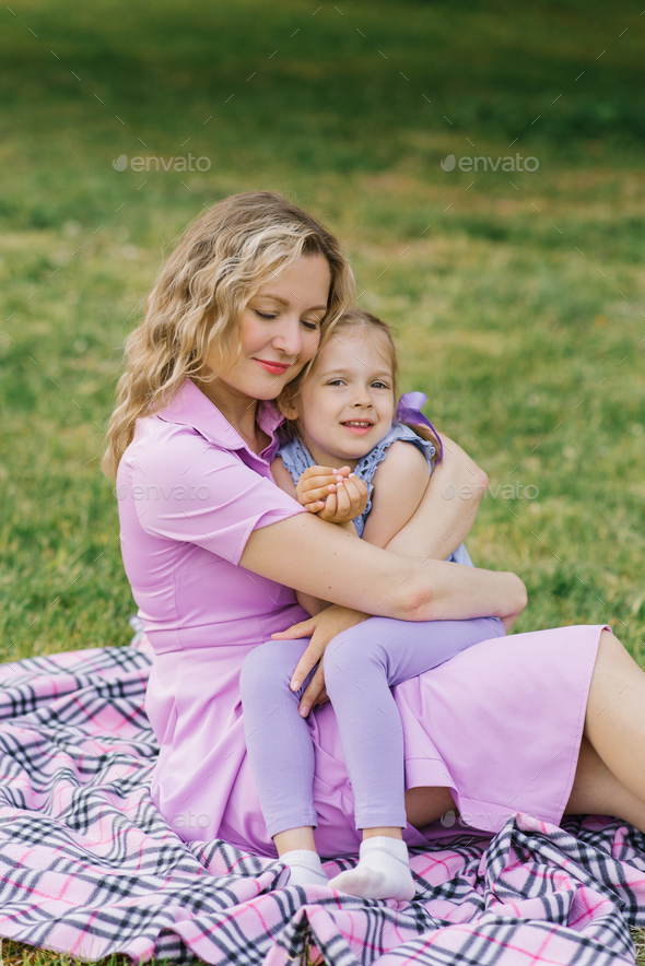 Mom hugs her little daughter in the park, sitting on a blanket Stock Photo by LeylaCamomile