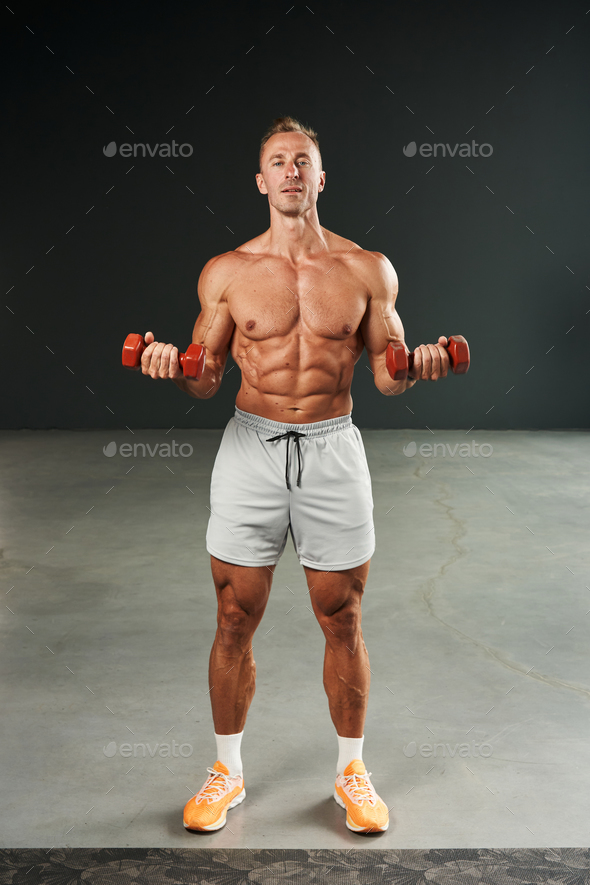 a handsome tanned caucasian athlete in shorts shakes his biceps with a ...