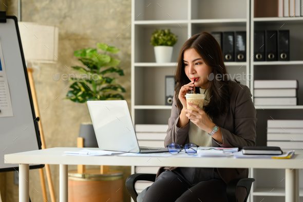 Pretty young employee holding glass of iced coffee and using laptop ...