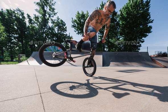 A mature shirtless guy with tattoos is balancing on his bmx in a skate ...