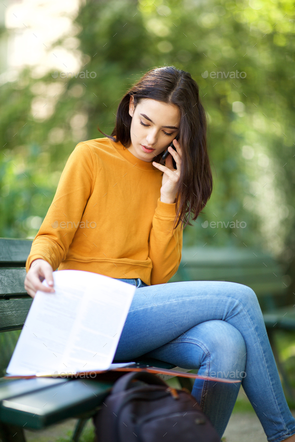 female university student sitting on park bench with notepad and mobile ...