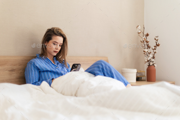 Young woman lying on a bed and scrolling her smartphone. Stock Photo by halfpoint