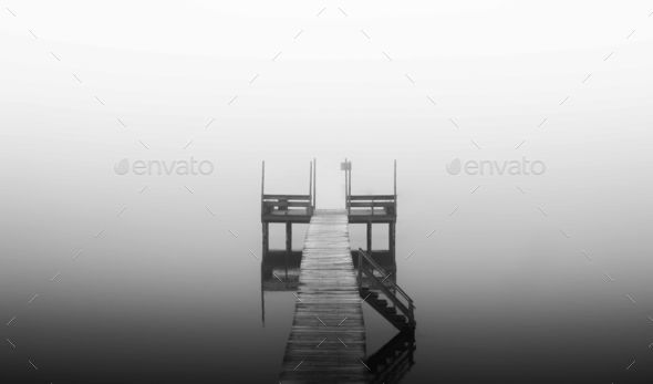 Grayscale of a serene dock shrouded in fog leading to the endless sea ...