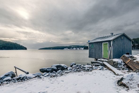 Winter scene of a small wooden boat house on a snowy shoreline beside a ...