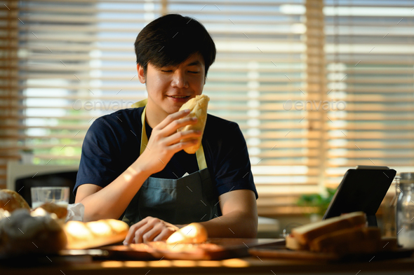 Millennial asian man holding fresh bread and using laptop at dining ...