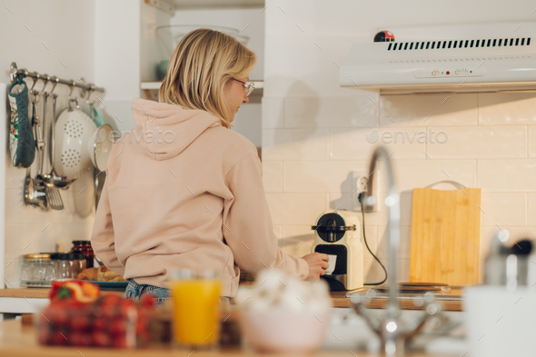 Back view of a blond woman standing in the kitchen and making coffee on ...