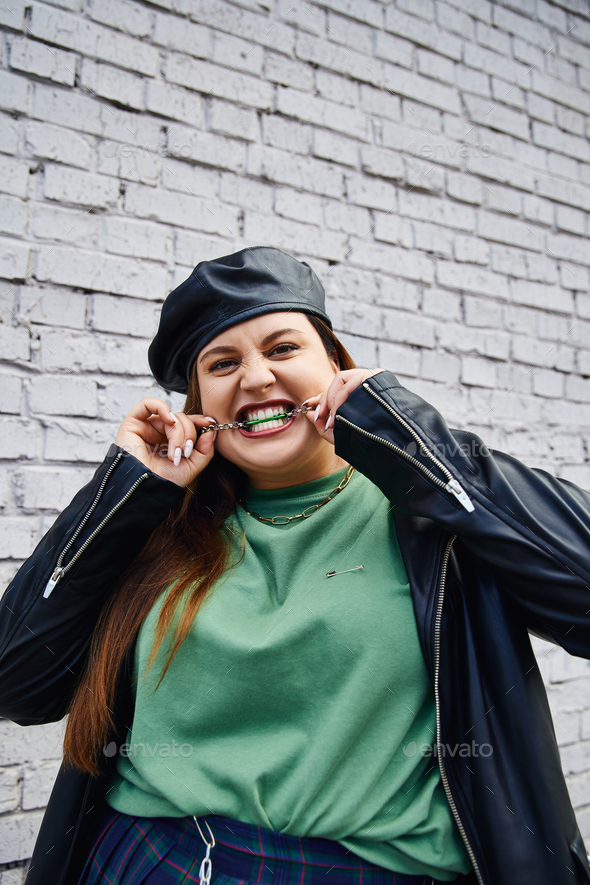 stylish plus size woman in leather jacket and black beret biting chain ...
