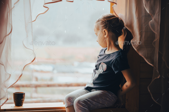 Sad child looking through rainy window at home Stock Photo by ninelutsk