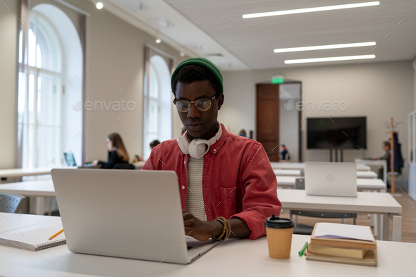 Puzzled student black hipster man sits at table with textbooks laptop ...