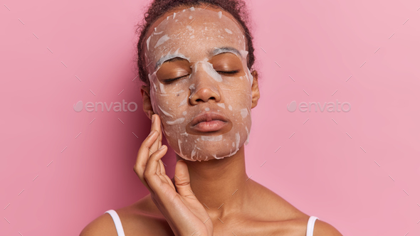 Indoor shot of dark skinned young woman with cloth moisturizing mask ...