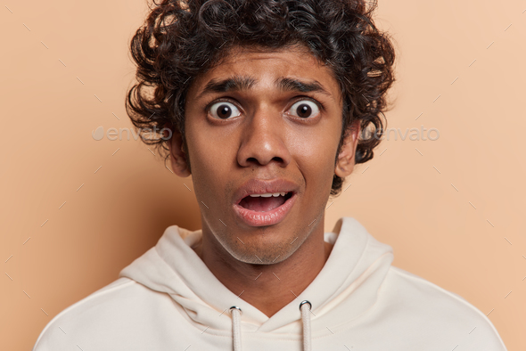 Close up shot of embarrassed curly haired man stares in disbelief ...