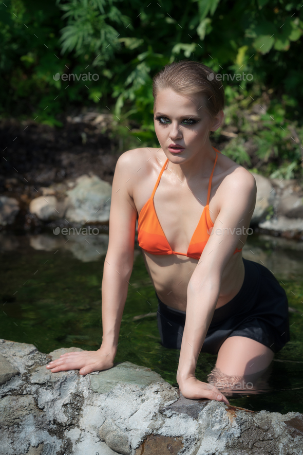 Woman stands in water of outdoor pool with geothermal water, leaning sensually on rocky edge of ...