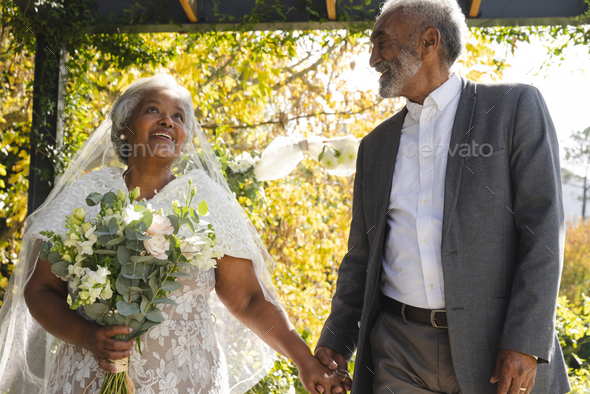 Happy senior biracial bride and groom holding hands at sunny outdoor ...