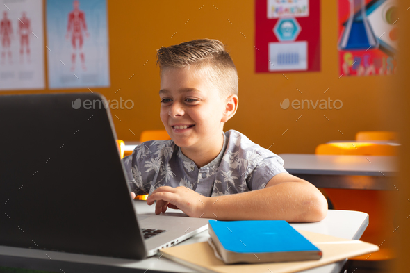Caucasian boy sitting at a desk in classroom using laptop and smiling ...