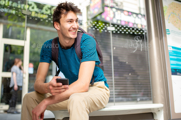 Close up smiling young man at bus stop holding mobile phone and looking ...