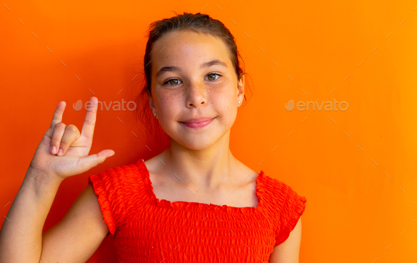 Happy caucasian schoolgirl doing sign language with hand over orange ...