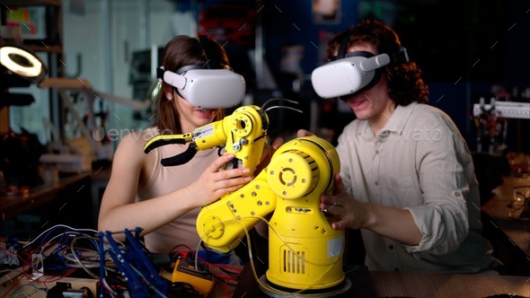Two young engineers fixing a yellow robot arm in the workshop, wearing ...