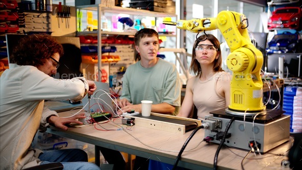 Three young happy engineers fixing an yellow robotic arm in the ...