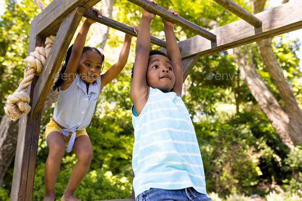 Low angle view of african american sister and brother hanging on monkeys bars at playground ...