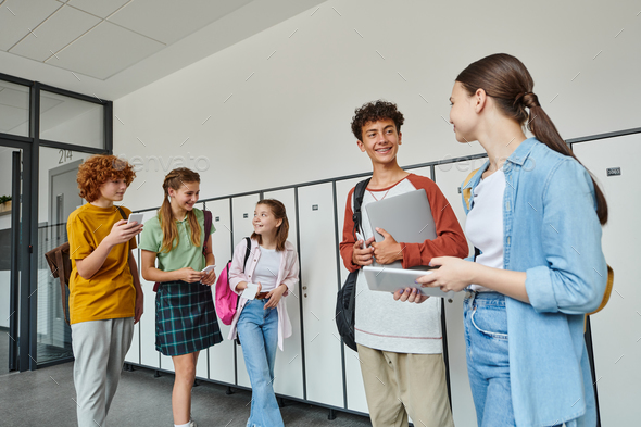 happy classmates talking and holding devices in hallway, back to school ...