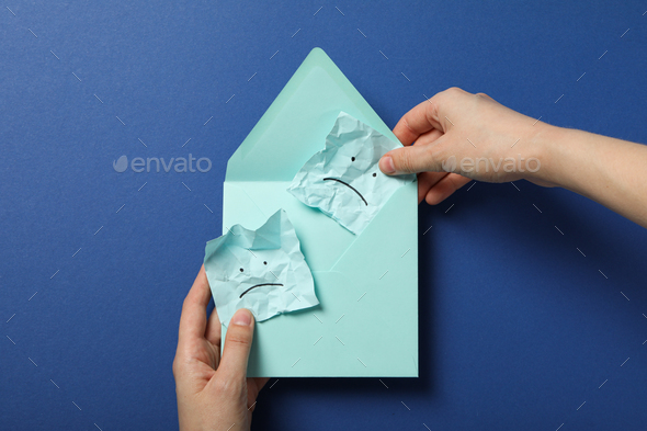 Envelope with papers with sad emoji in hands on blue background, top ...