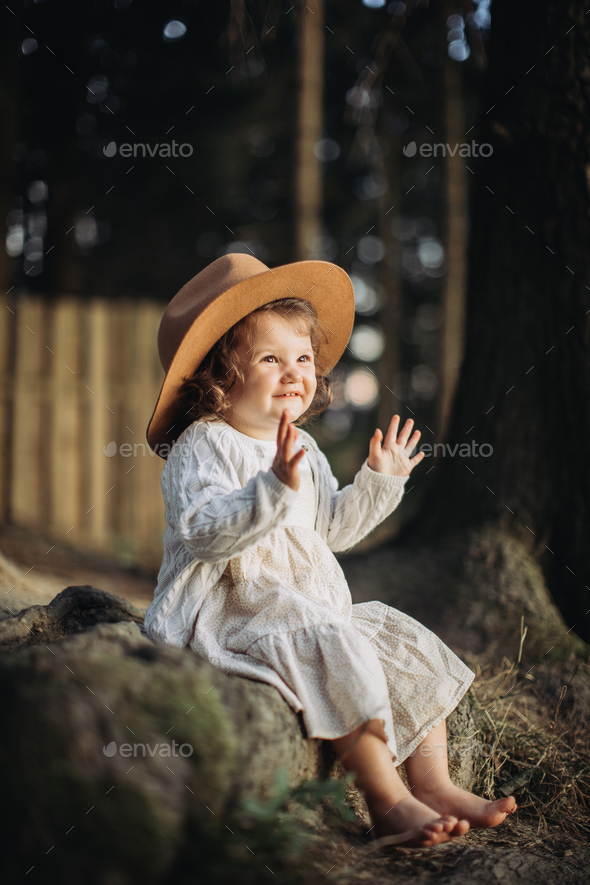 Excited little girl wearing hat, sitting bootless in the countryside ...