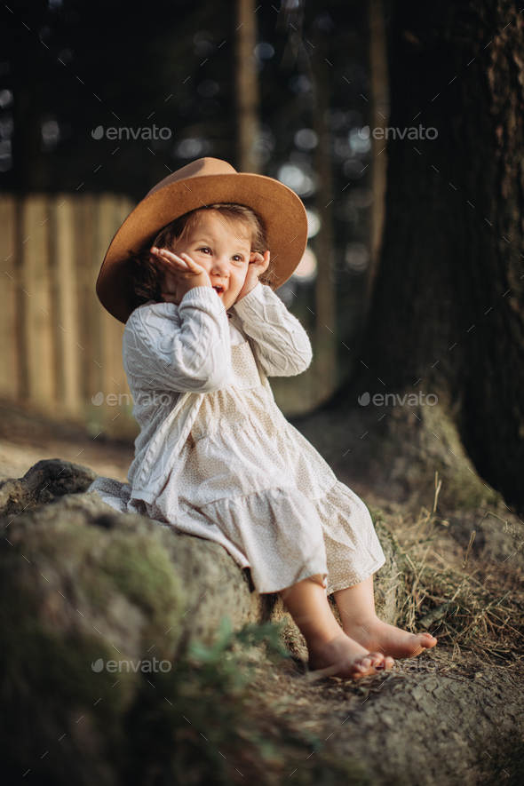 Excited little girl wearing hat, sitting bootless in the countryside ...