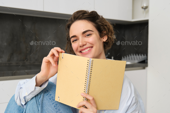 Image of brunette girl at home, holding notebook, reading through her ...