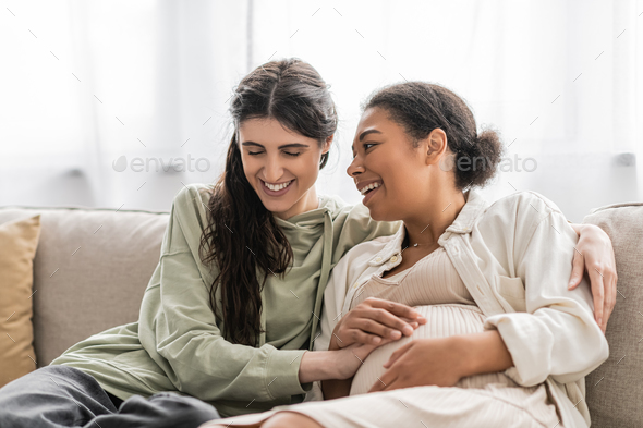 Overjoyed Lesbian Woman Hugging Pregnant Multiracial Wife While Sitting