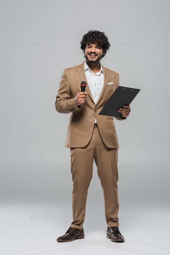 Full length of smiling indian event host in brown formal wear holding ...