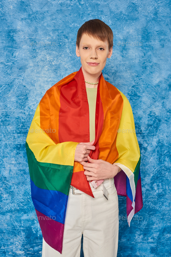 Portrait of young queer person wrapped in lgbt flag looking at camera ...