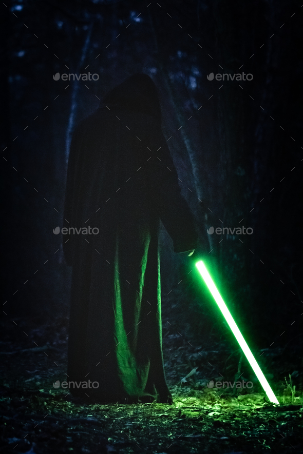 Man in a black mantle holding a green neon sword in the hand. Dark ...