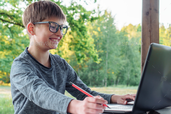 Boy with glasses sit at park table, look at laptop screen and smiling ...