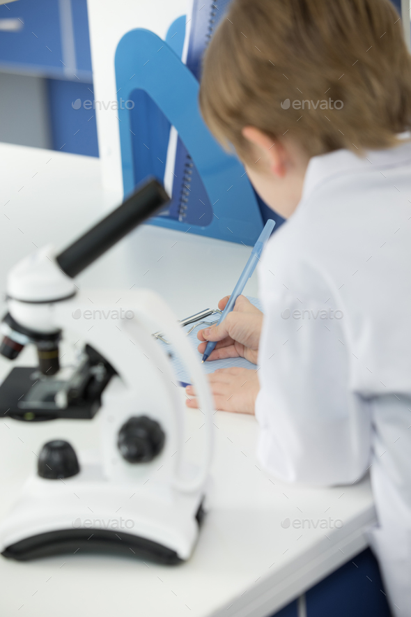 Schoolboy in white lab coat taking notes during scientific experiment ...
