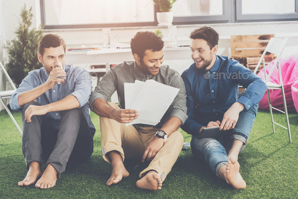 Smiling young barefoot businessmen sitting with papers and using digital tablet, business ...