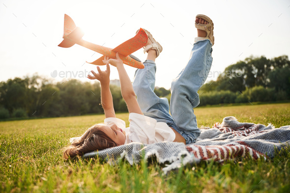 Laying down on the ground. Happy little girl is playing with toy plane ...