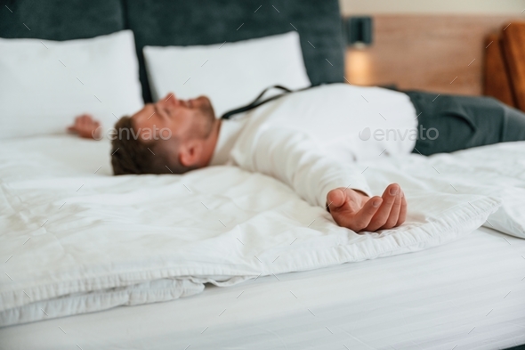 Tired, lying down on the bed. Businessman is indoors in the hotel room ...