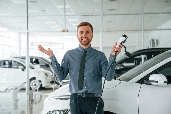 Smiling, holding the charger. Handsome car dealership worker is with ...