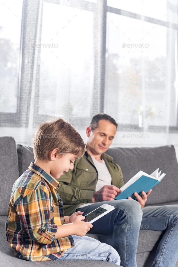 handsome father reading book while little son using tablet on couch ...