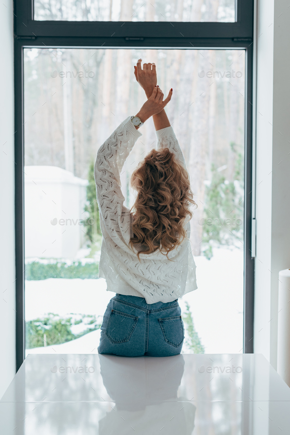 Back view of curly woman standing with hands up in front of window ...