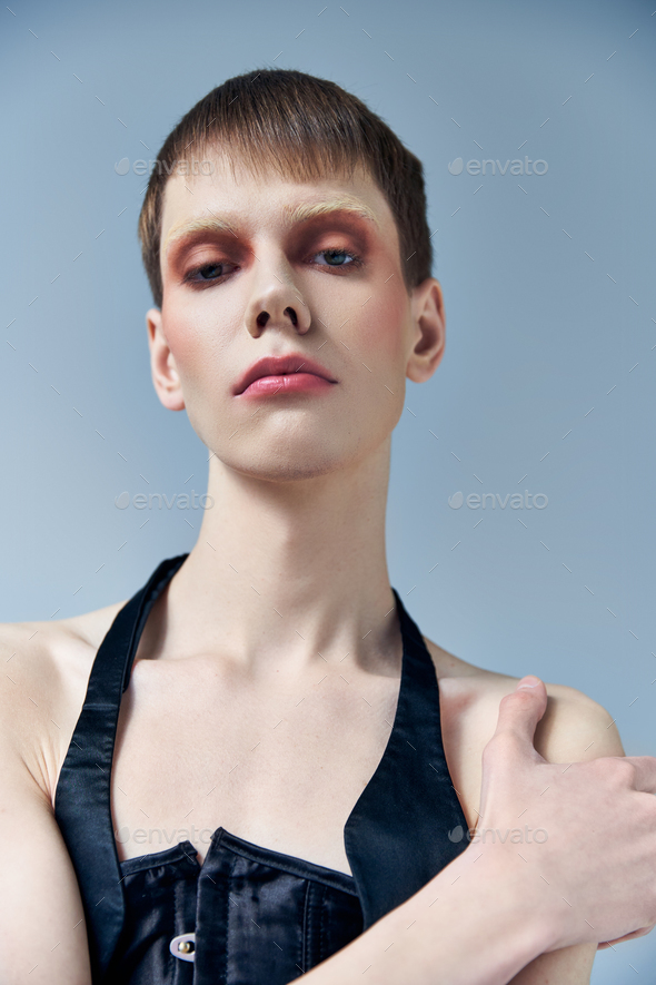 portrait, queer model with makeup looking at camera, posing on grey ...
