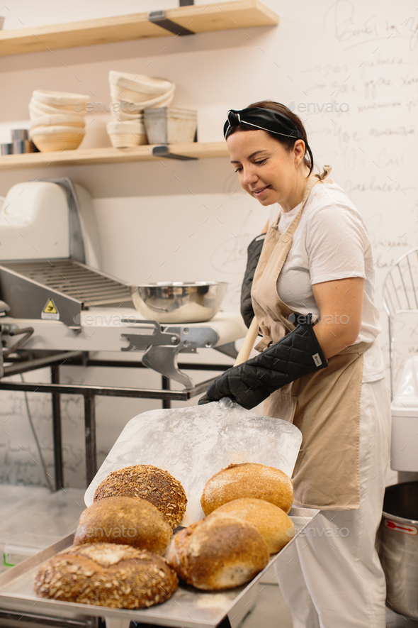 Female baker using a peel to take out a loaf of bread of the oven in a ...