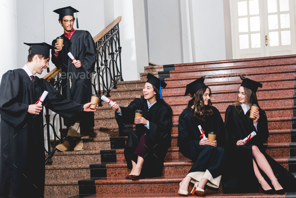 cheerful group of students in graduation gowns sitting on stairs with ...