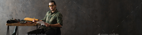 Bearded focused craftsman writing down notes while working with leather ...